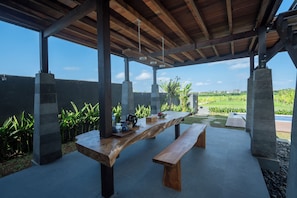 Um pátio externo com mesa e bancos de madeira, rodeado por vegetação e uma vista aberta do céu.