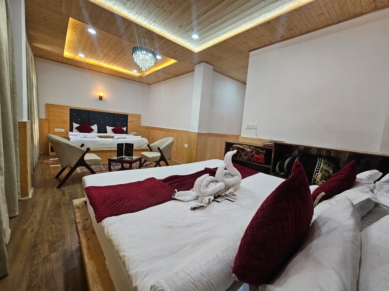 Quarto de hotel com teto de madeira, um lustre, cama com edredom branco e almofadas vermelhas, cabeceira de madeira e uma pequena mesa com uma cadeira.
