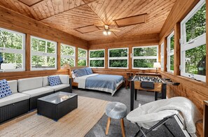 Interior acolhedor de cabana com sofá, cama e mesa de futebol, grandes janelas com vista para o verde.