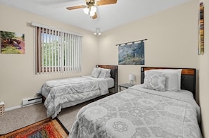Quarto de hotel com duas camas, ventilador de teto, janela com persianas e quadros nas paredes.
