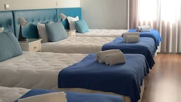 Quarto de hotel com várias camas dispostas em fila, cada uma com colcha azul e almofadas brancas.