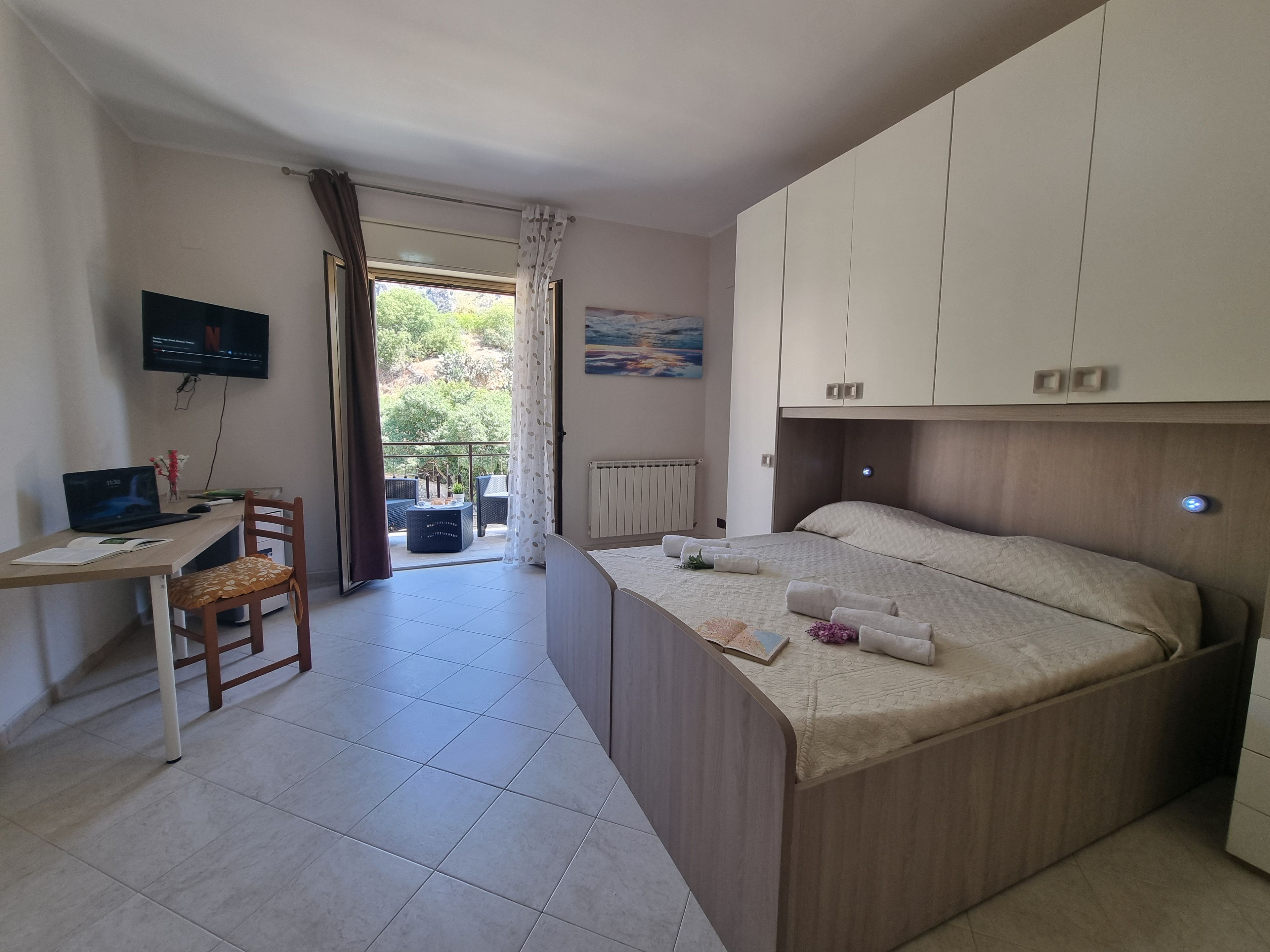 Quarto com cama, escrivaninha com laptop, cadeira e vista para o exterior.