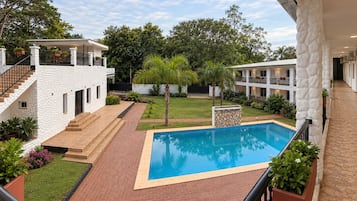 Uma área de piscina com piscina azul límpida, rodeada por um pátio de tijolos, e adjacente a um edifício branco com varanda.