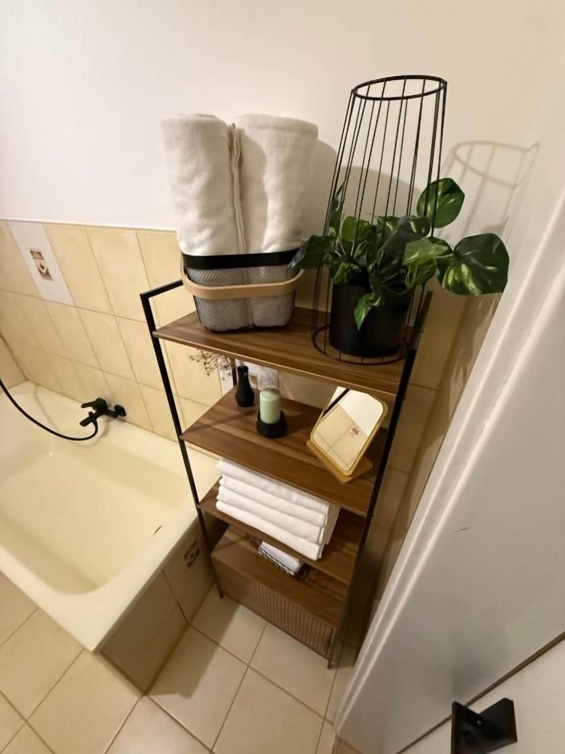 Um banheiro com banheira, prateleira com toalhas, planta em vaso e um suporte de metal.