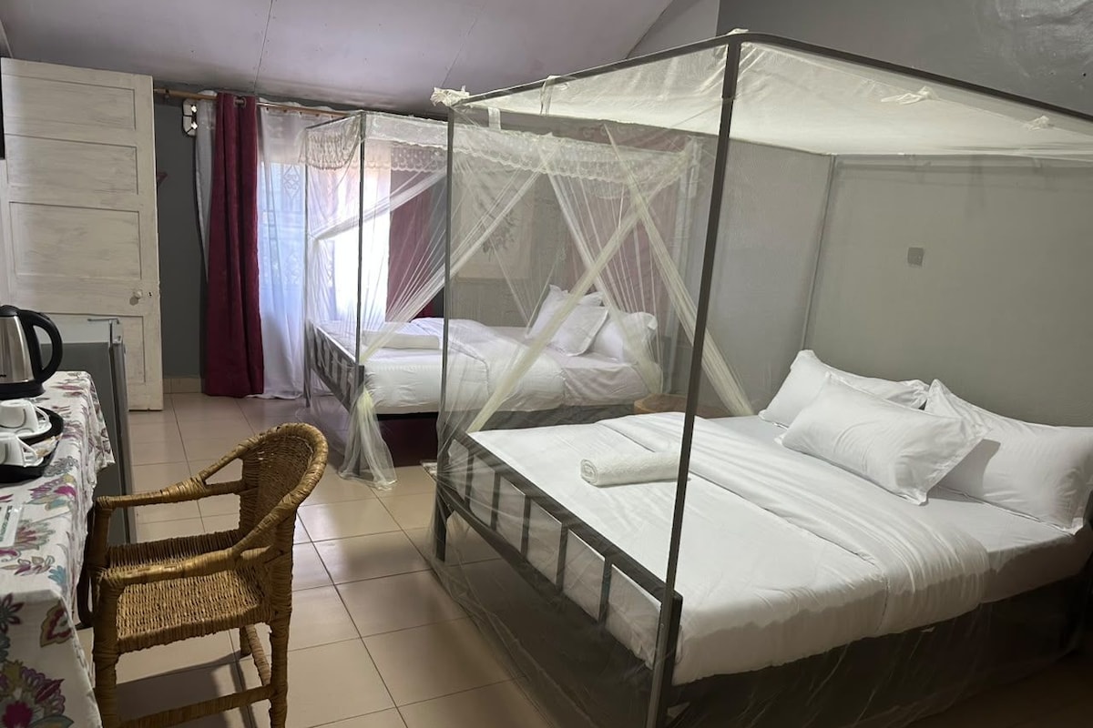 Quarto com cama de dossel, uma cadeira de vime, escrivaninha com chaleira e janela com cortinas.