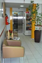 Um saguão de hotel com balcão de recepção, área de estar, geladeira abastecida com bebidas, extintor de incêndio e elevador.