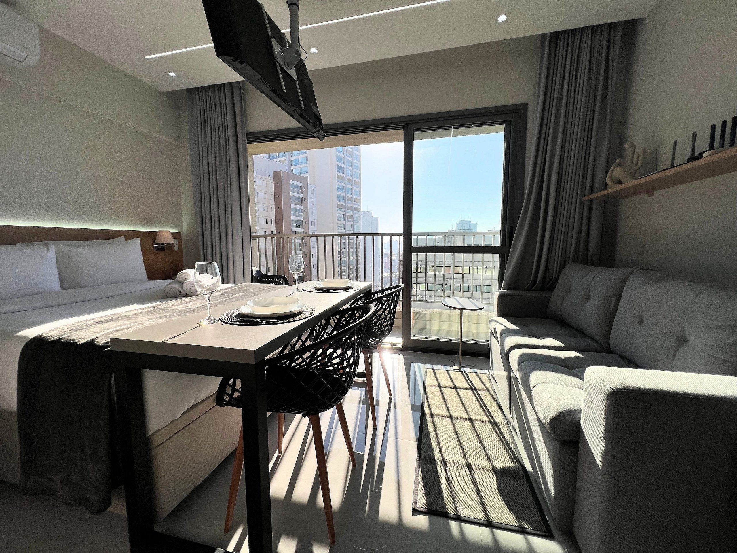 Quarto de hotel com cama, mesa de jantar com cadeiras, sofá e varanda com vista para edifícios.