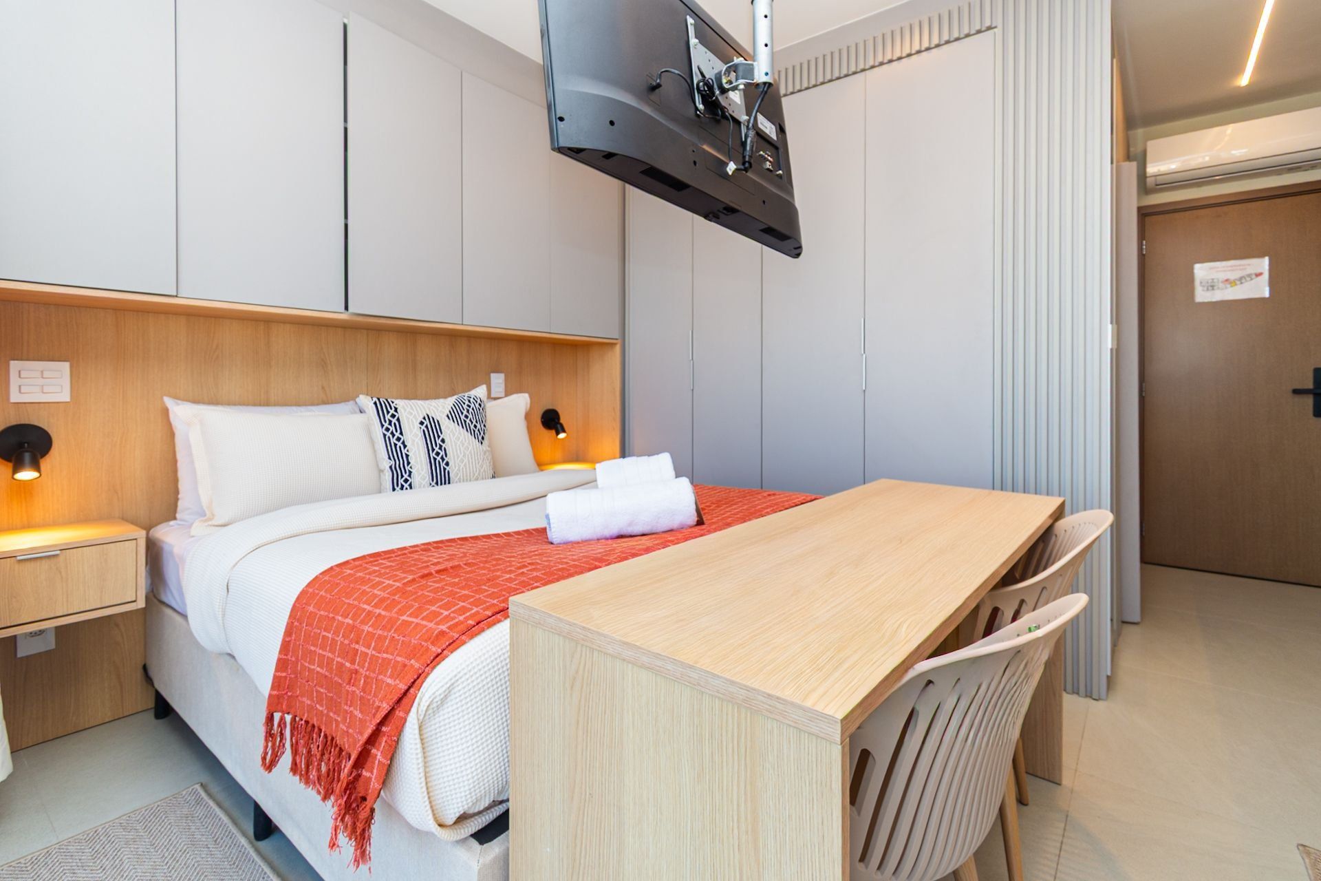 Quarto de hotel com cama, escrivaninha de madeira, cadeira, televisão na parede e porta.