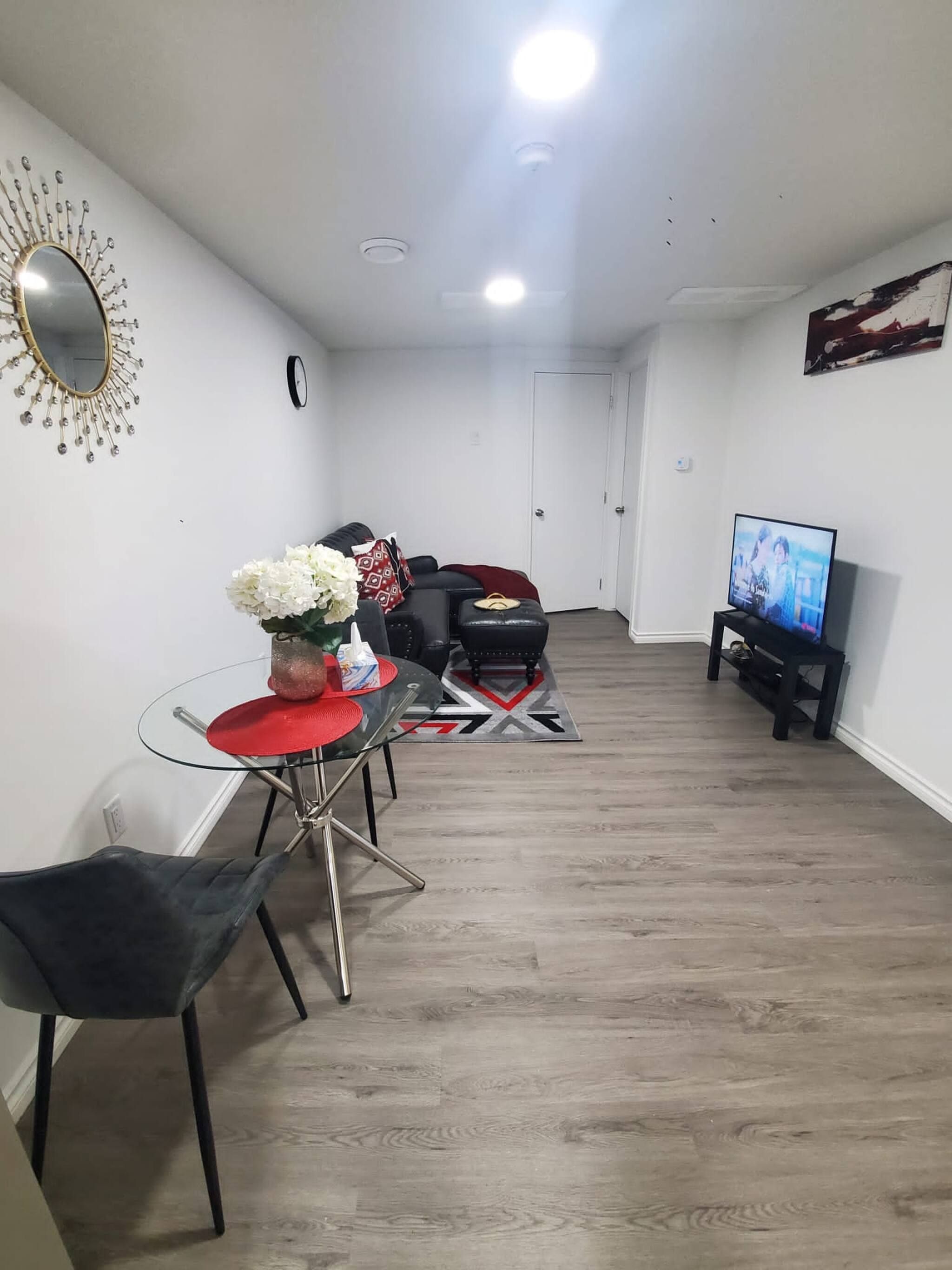Uma sala de estar moderna com uma mesa de jantar de vidro, cadeiras pretas, sofá, televisão e um espelho decorativo na parede.