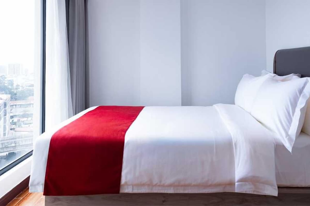 Quarto de hotel com cama, janela com cortinas e vista para prédios.