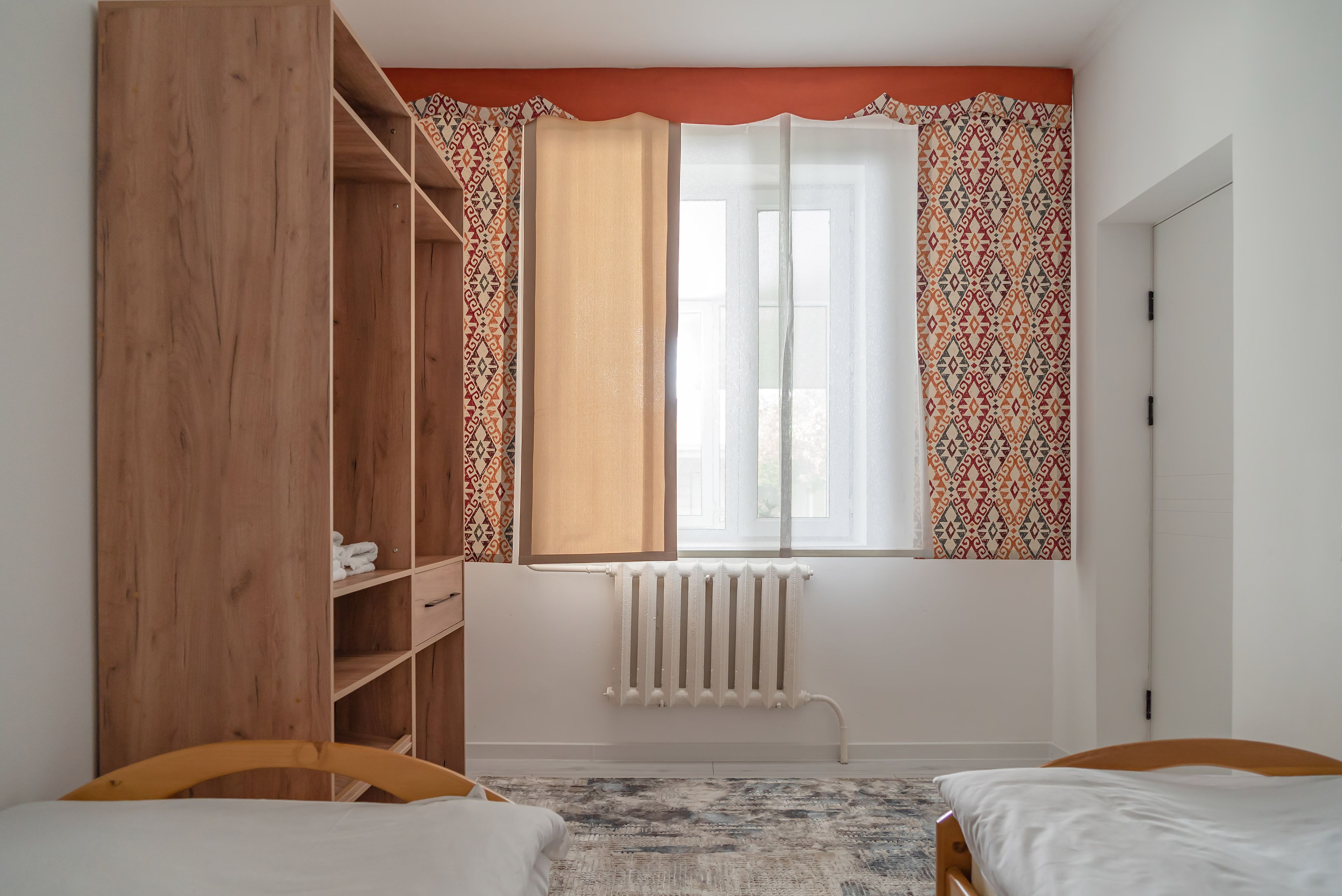 Quarto com guarda-roupa de madeira, radiador, janela com cortinas estampadas e cama.