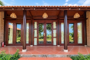 Uma área externa coberta, com pilares de madeira, grandes janelas e piso de azulejos.