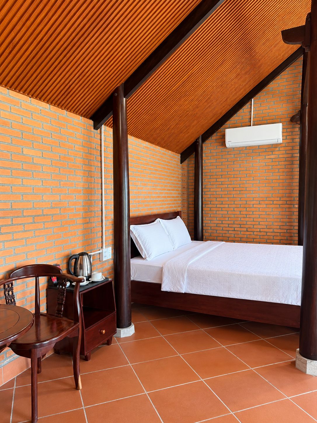 Uma cama de solteiro com cabeceira de madeira, mesa de cabeceira com chaleira e uma cadeira em um quarto com paredes de tijolo e teto inclinado.