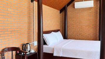 Uma cama de solteiro com cabeceira de madeira, mesa de cabeceira com chaleira e uma cadeira em um quarto com paredes de tijolo e teto inclinado.