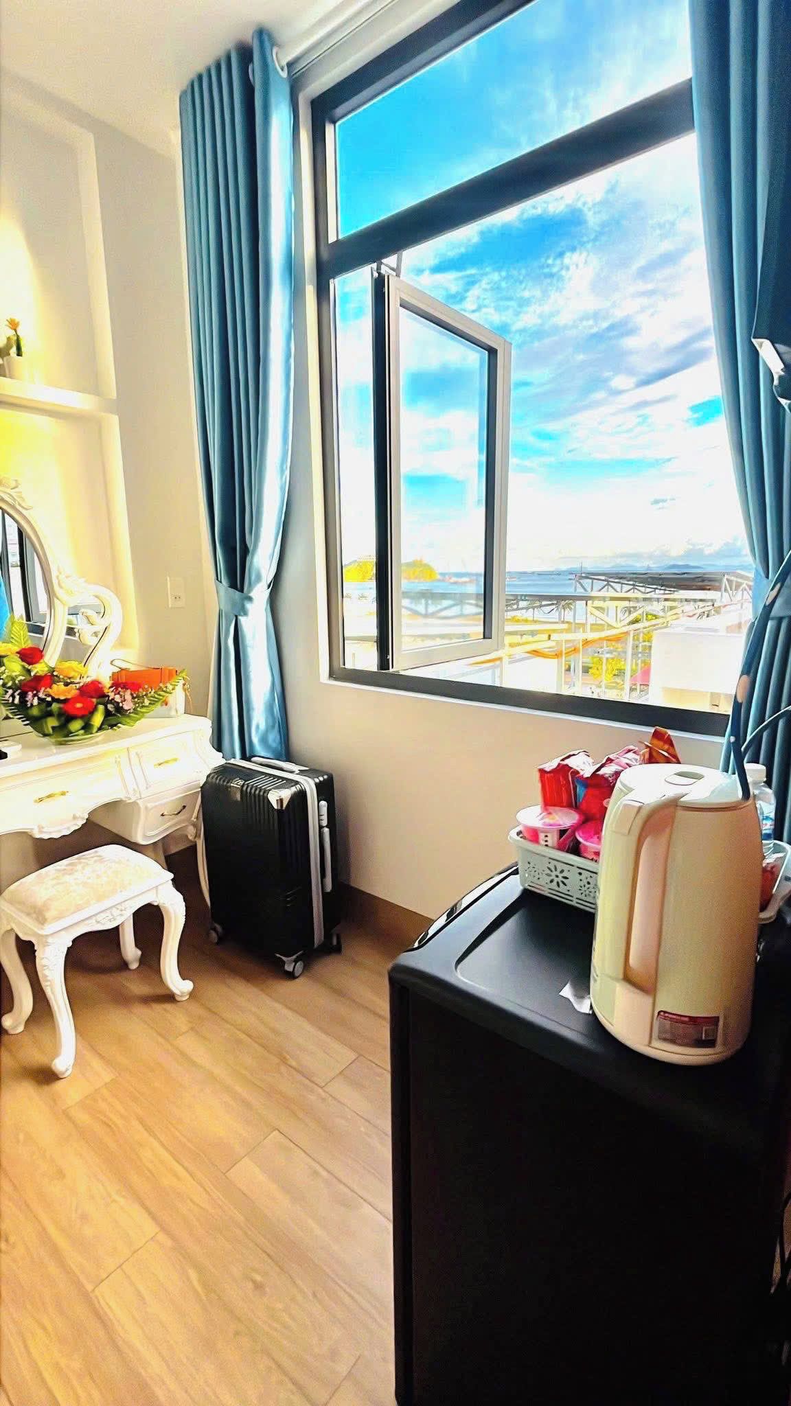 Quarto de hotel com uma grande janela, uma escrivaninha com abajur, uma mala e um banquinho pequeno.