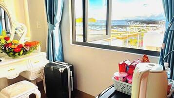 Quarto de hotel com uma grande janela, uma escrivaninha com abajur, uma mala e um banquinho pequeno.