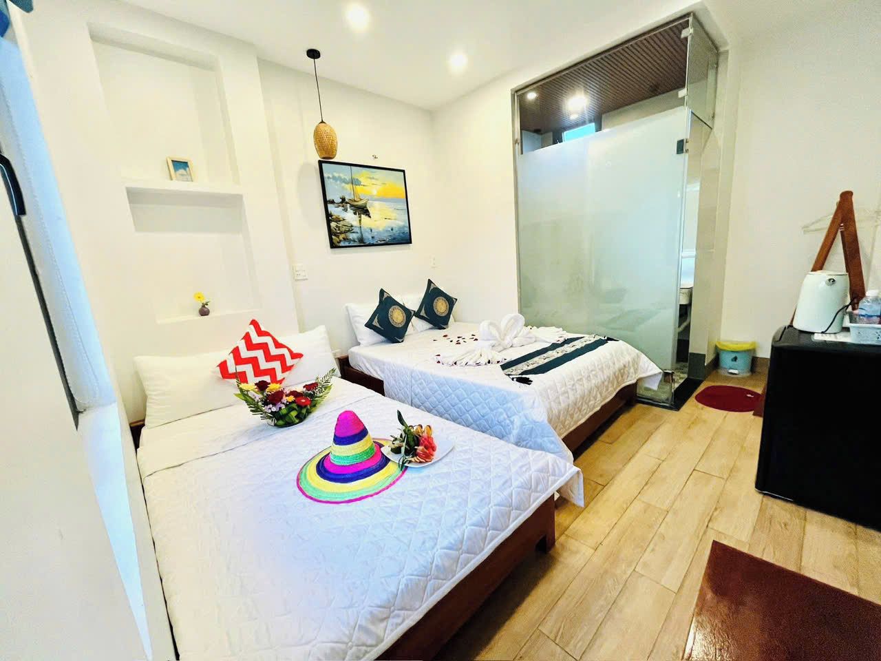 Quarto de hotel com duas camas, uma escrivaninha, uma cadeira, uma mesa pequena, um quadro na parede e uma janela.
