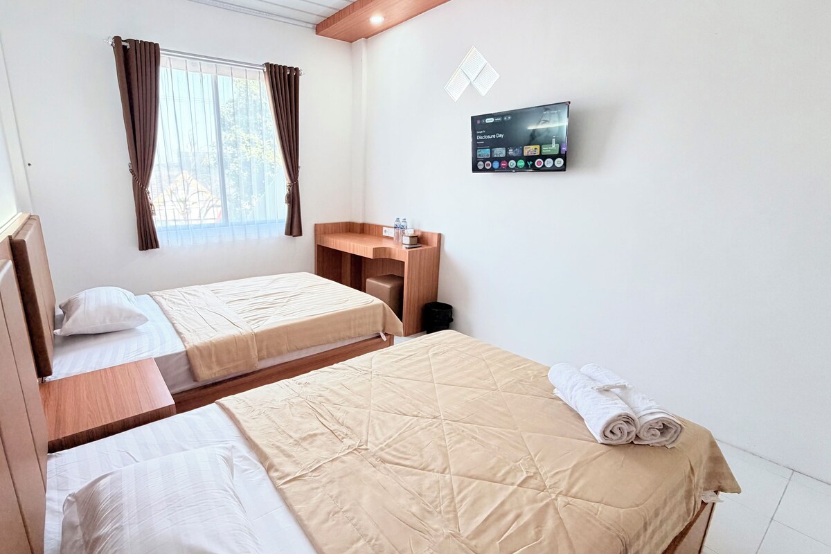 Quarto de hotel com duas camas, uma escrivaninha, janela com cortinas e painel de controle na parede.