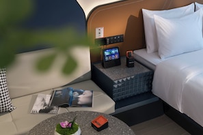 Quarto de hotel moderno com cama, uma mesinha com uma revista e uma flor, rack para TV com controle remoto e tomada na parede.