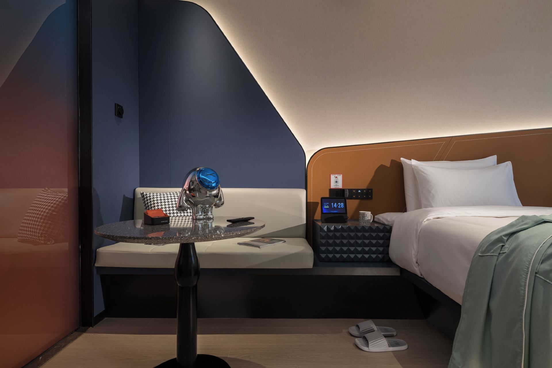 Quarto de hotel com cama, mesa de cabeceira com relógio, pequena mesa com um objeto decorativo e um sofá.