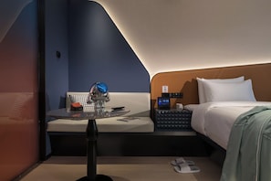 Quarto de hotel com cama, mesa de cabeceira com relógio, pequena mesa com um objeto decorativo e um sofá.