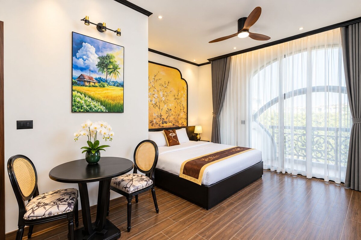 Quarto de hotel com cama, mesa de jantar e cadeiras, ventilador de teto e uma grande janela com cortinas.