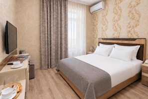Quarto de hotel com cama, televisão, escrivaninha com xícara e pires, e janela com cortinas.