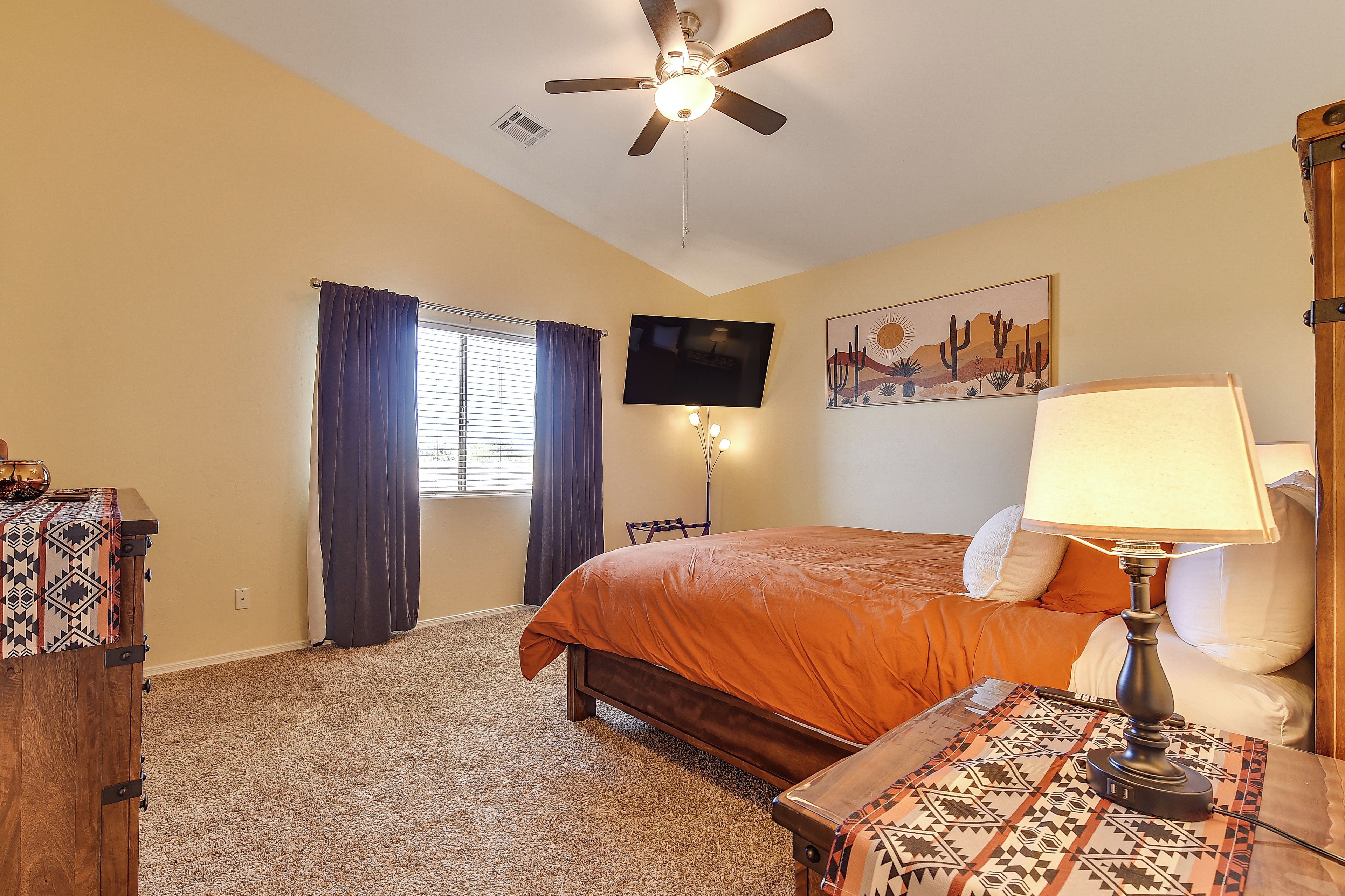 Quarto de hotel com cama, televisão, ventoinha no teto, quadro e luminária.