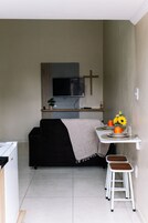 Um espaço de estar pequeno e moderno, com uma TV de tela plana na parede, um sofá, uma mesa de jantar com um vaso de girassóis e um banquinho.