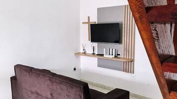 Quarto com sofá marrom escuro, TV de tela plana presa a uma prateleira de madeira e escada ao fundo.