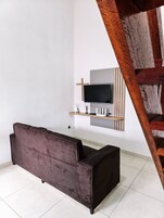 Quarto com sofá marrom escuro, TV de tela plana presa a uma prateleira de madeira e escada ao fundo.