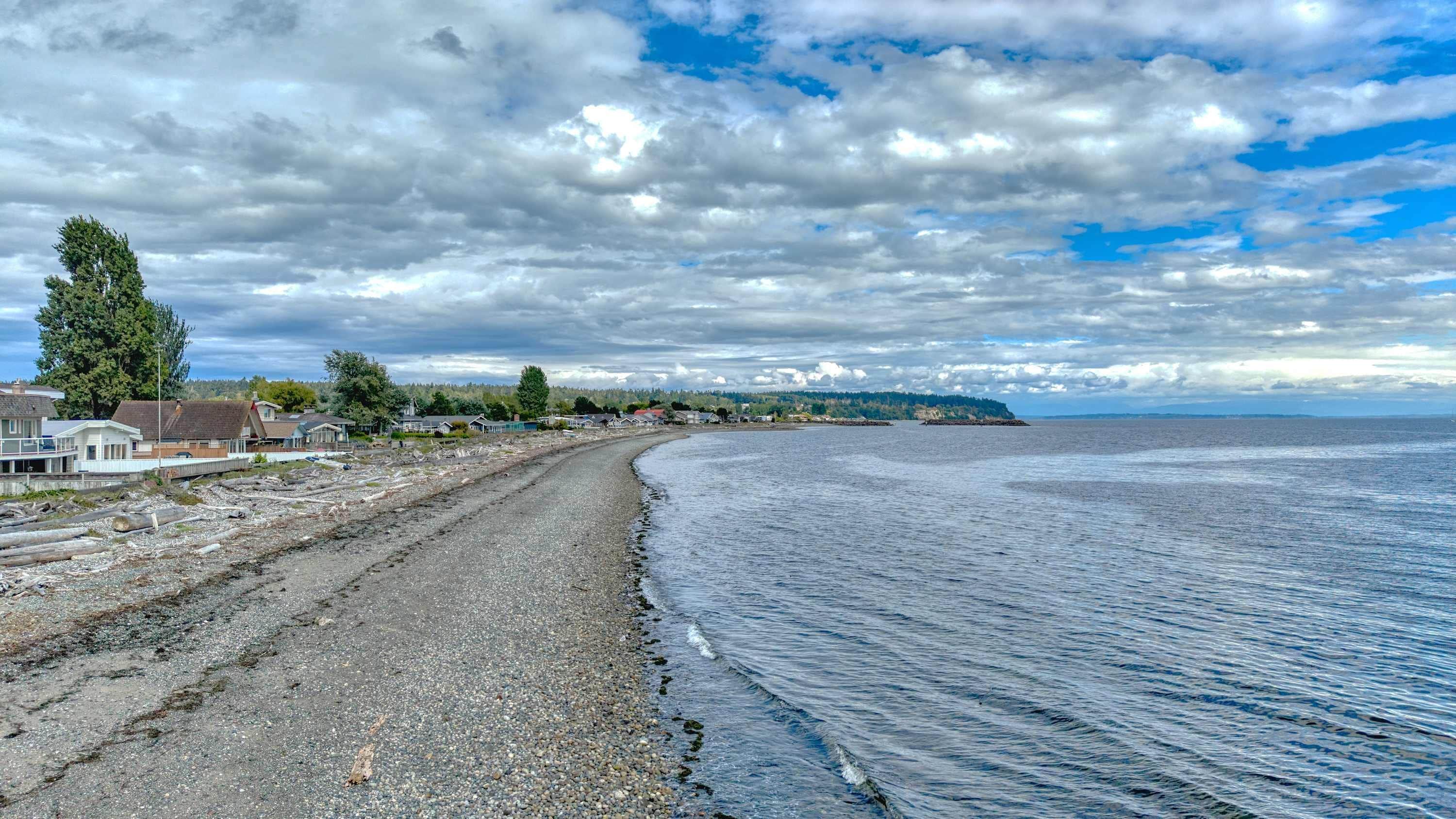 Uma estrada costeira com casas à esquerda, que leva a uma praia com águas calmas e céu nublado.