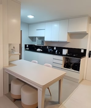 Cozinha moderna com armários brancos, bancada preta e área de jantar com mesa e cadeiras brancas.