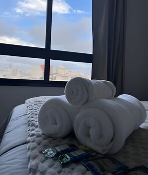 Cama com toalhas brancas dobradas e janela com vista para a cidade.