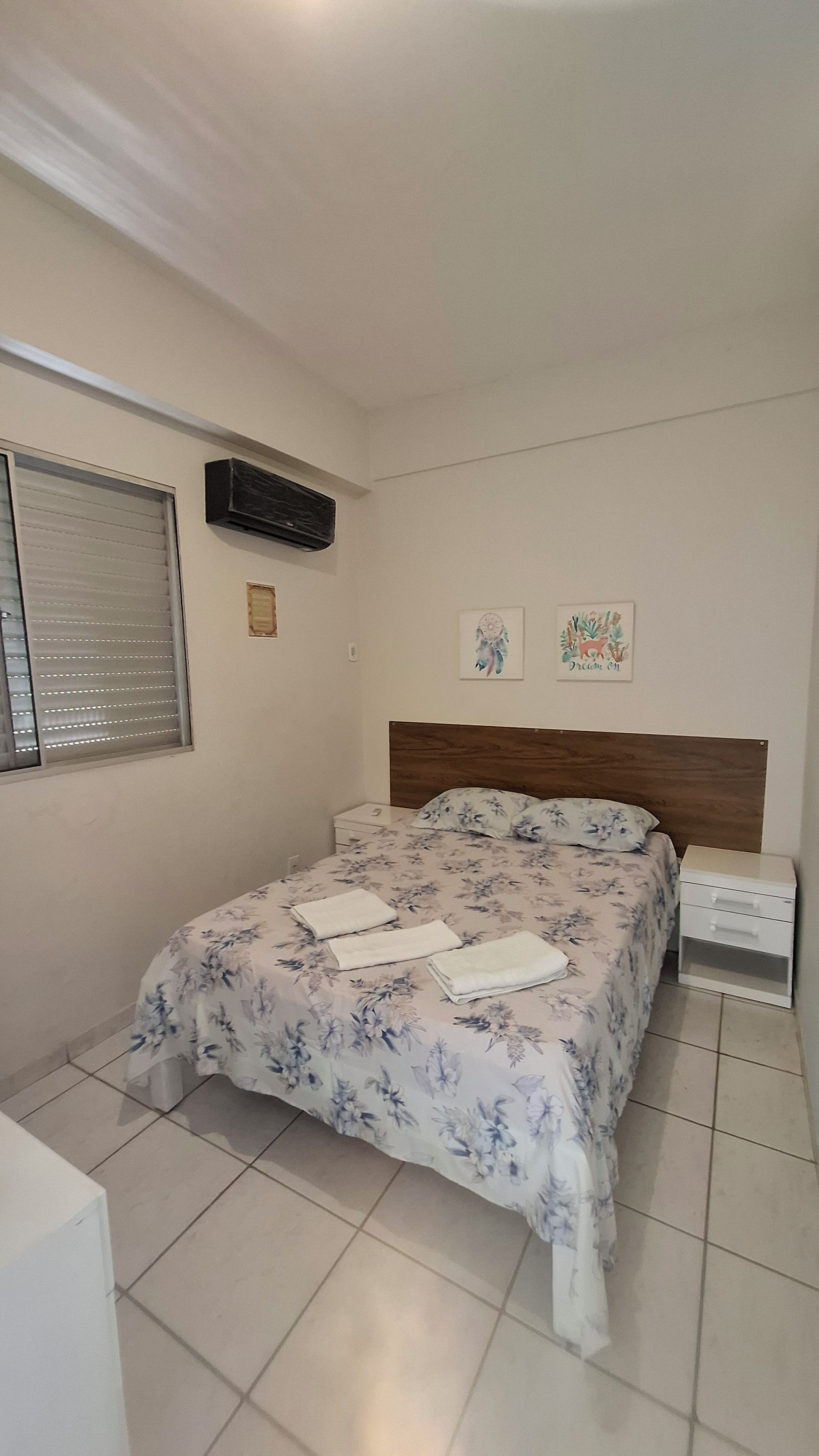 Quarto com cama, mesa de cabeceira, janela com persianas, ar condicionado e duas obras de arte penduradas na parede.