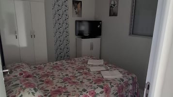 Quarto com cama, guarda-roupa, televisão sobre um suporte e uma janela.