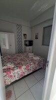 Quarto com cama, guarda-roupa, televisão sobre um suporte e uma janela.
