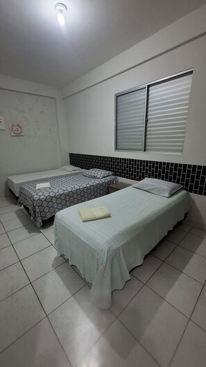 Quarto com três camas, janela com persianas, piso ladrilhado e luminária fixada na parede.