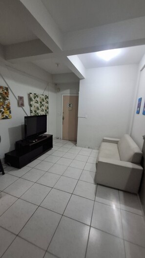 Uma sala de estar com televisão de tela plana em um suporte, um sofá bege e piso revestido de azulejos.