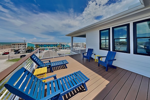 Um terraço com cadeiras de descanso azuis e amarelas, uma mesa pequena e vista para a praia e edifícios ao fundo.
