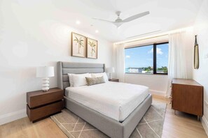 Quarto com cama, mesas de cabeceira, cômoda, ventilador de teto e uma grande janela com vista.