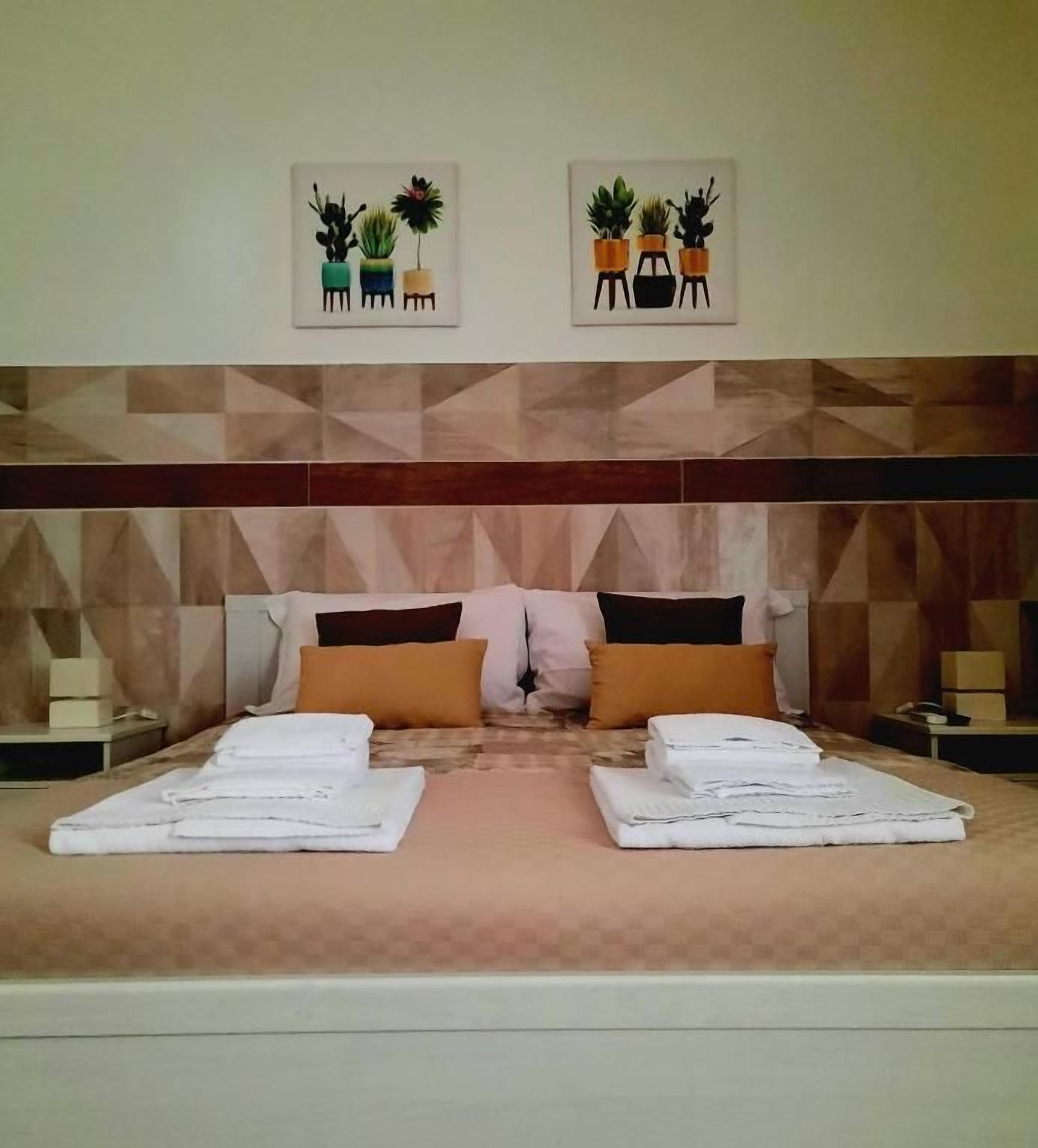 Quarto de hotel com uma cama, duas mesas de cabeceira, um painel de cabeceira com padrão geométrico e duas obras de arte na parede retratando plantas em vasos.