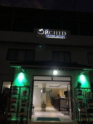 Exterior do The Orchid Grand Hotel, com uma placa acima da entrada, plantas em vasos de ambos os lados e uma escadaria que leva ao saguão.