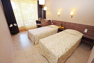Quarto de hotel com duas camas, uma escrivaninha, uma cadeira, uma mesinha de centro com um vaso de flores e uma janela com cortinas.