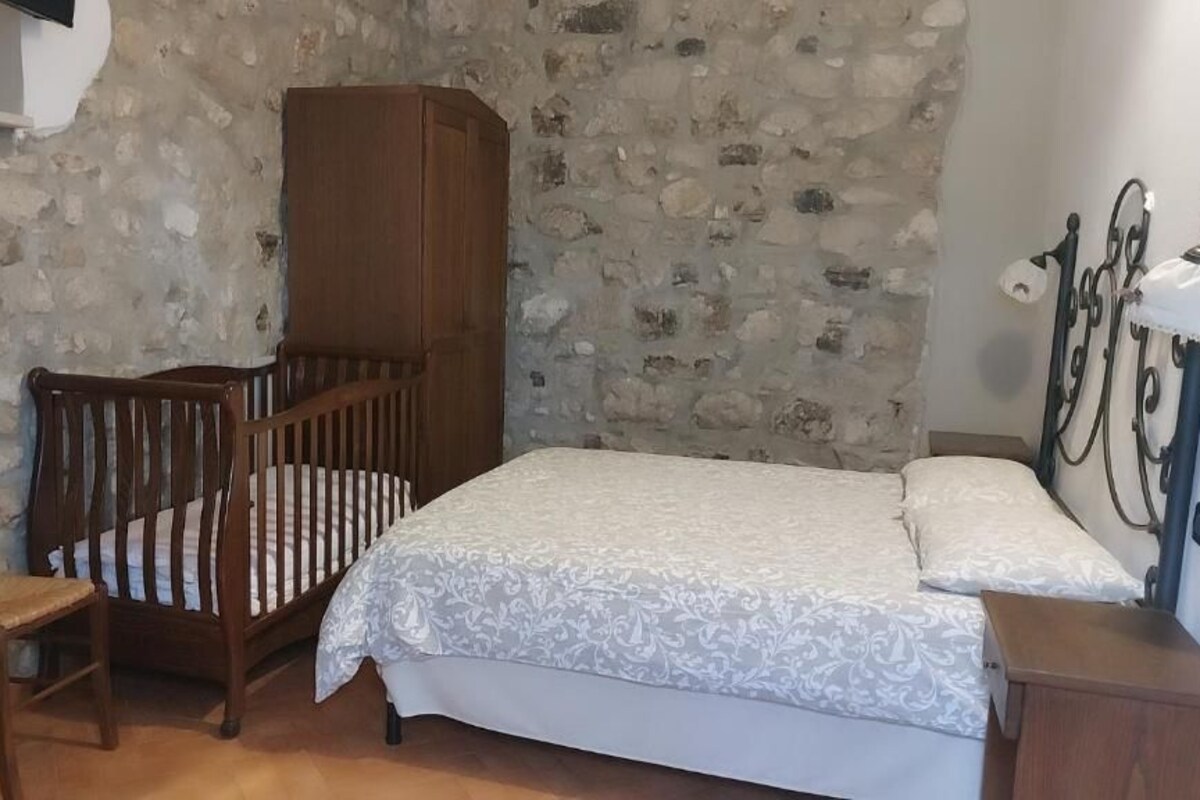 Quarto com parede de pedra, berço de madeira, cama com colcha estampada e mesa de cabeceira.