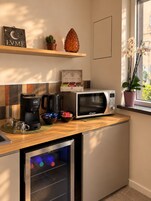 Uma cozinha compacta equipada com cafeteira, micro-ondas e um frigorífico pequeno. Há uma estante com uma planta em vaso e um objeto decorativo.