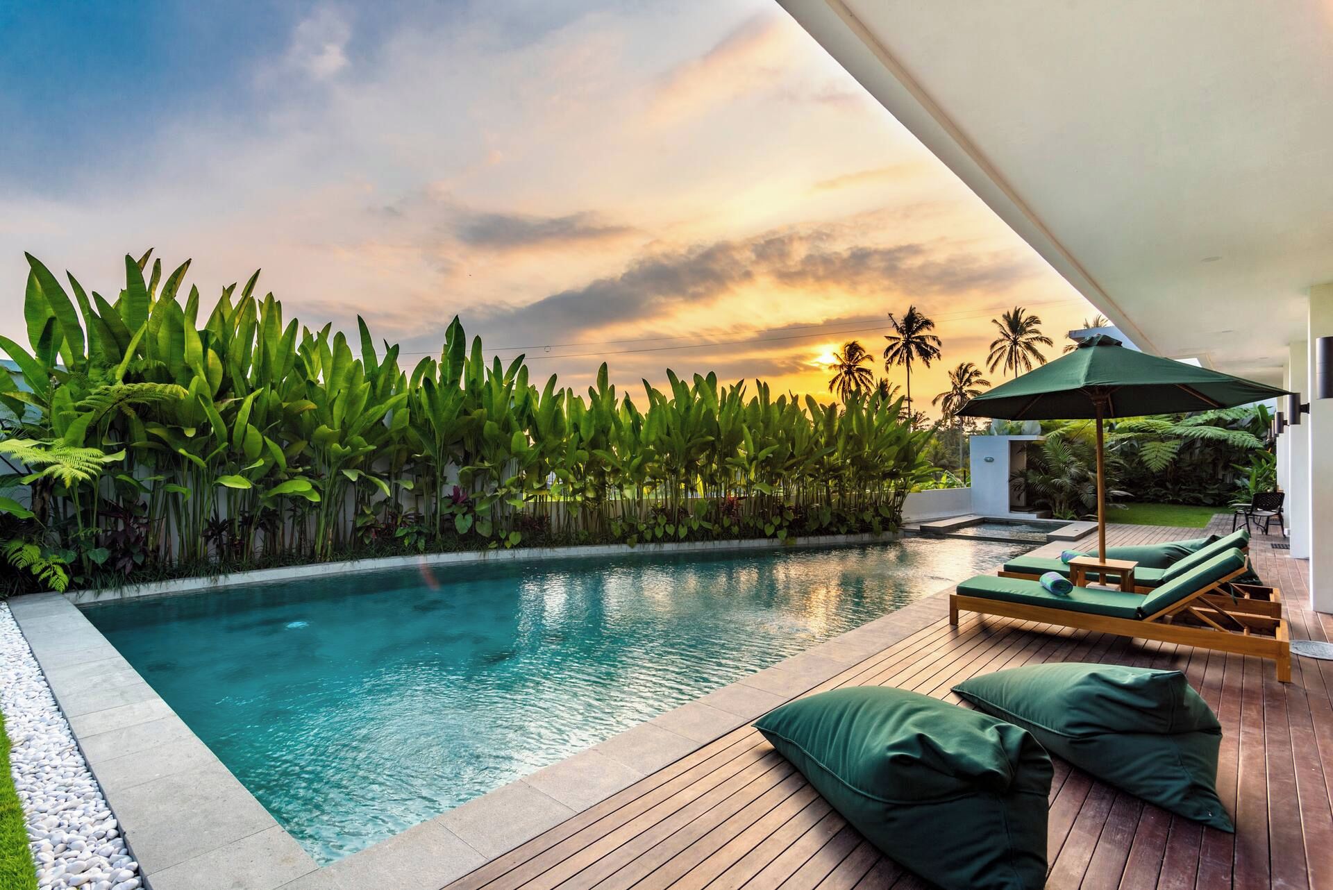 Área de piscina com espreguiçadeiras e guarda-sol, rodeada por plantas tropicais e palmeiras.