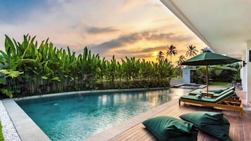 Área de piscina com espreguiçadeiras e guarda-sol, rodeada por plantas tropicais e palmeiras.