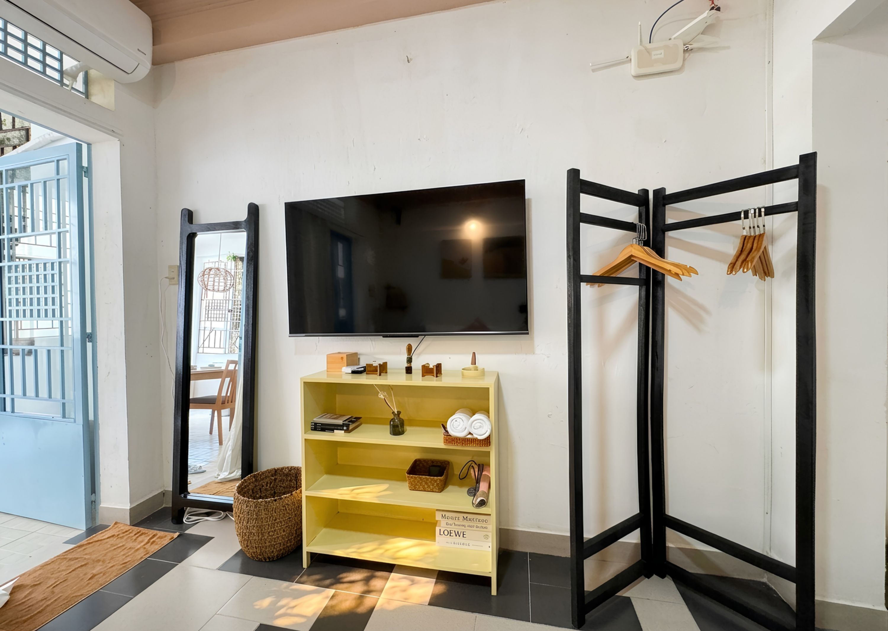 Quarto com televisão de tela plana fixada na parede, estante amarela com diversos objetos, guarda-roupa com moldura preta e cesta de vime no chão.