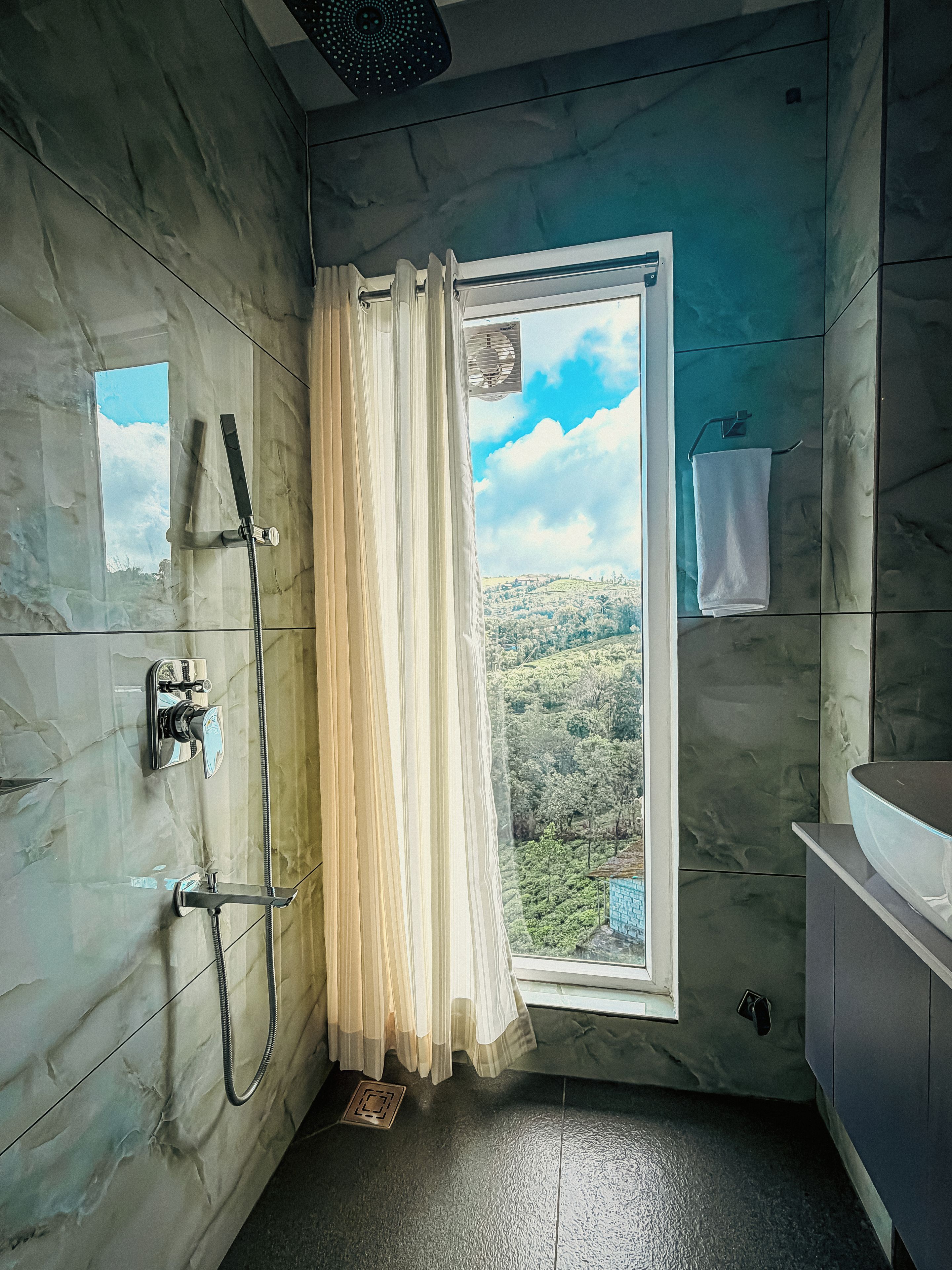 Casa de banho com chuveiro, janela com vista para paisagem verde e toalha pendurada na parede.